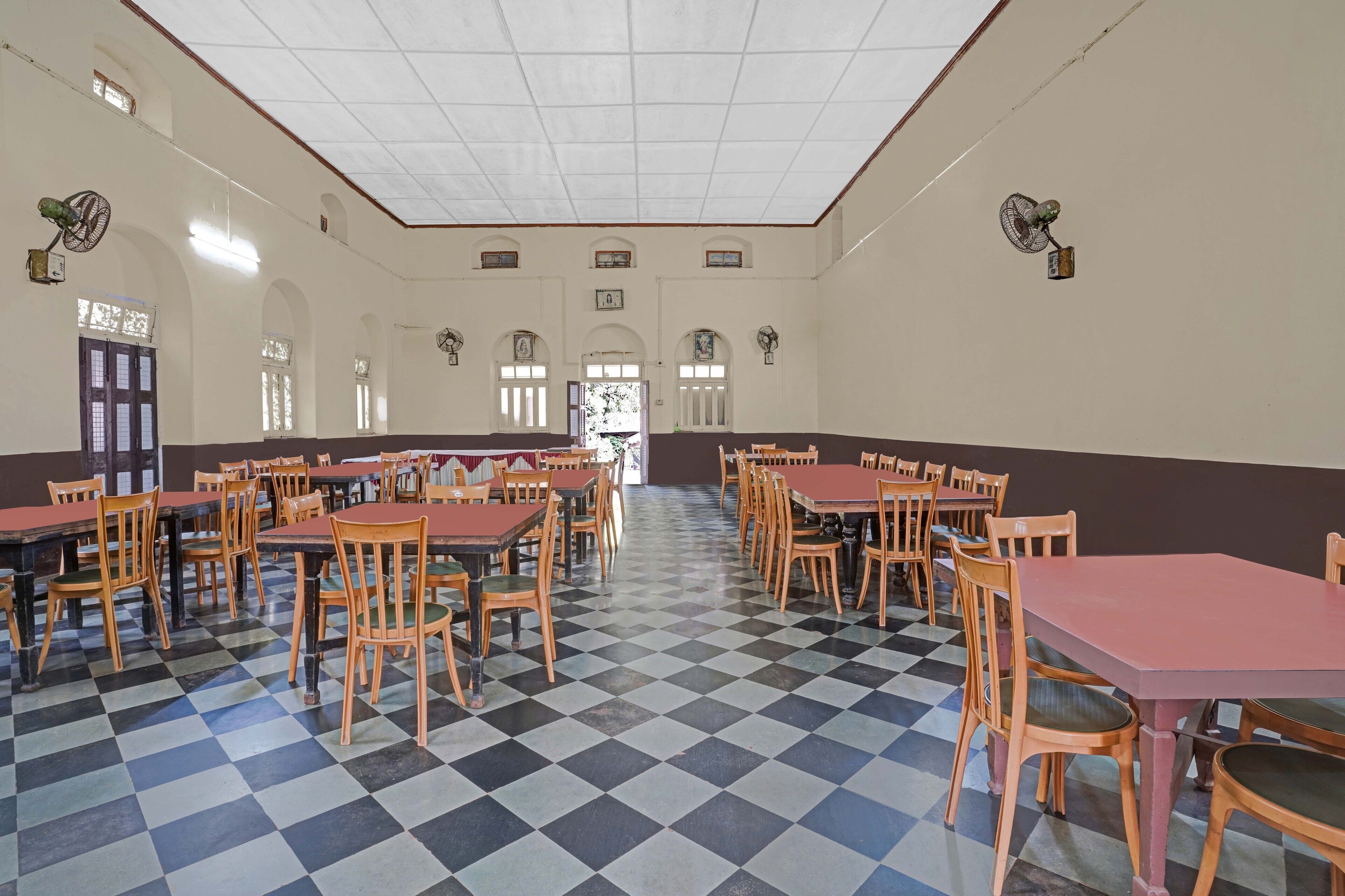 Uma sala de jantar ampla com piso xadrez, mesas e cadeiras de madeira e janelas em arco.