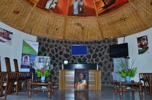 Uma área de recepção com parede de pedra, cadeiras de madeira, balcão de recepção e televisão fixada na parede.