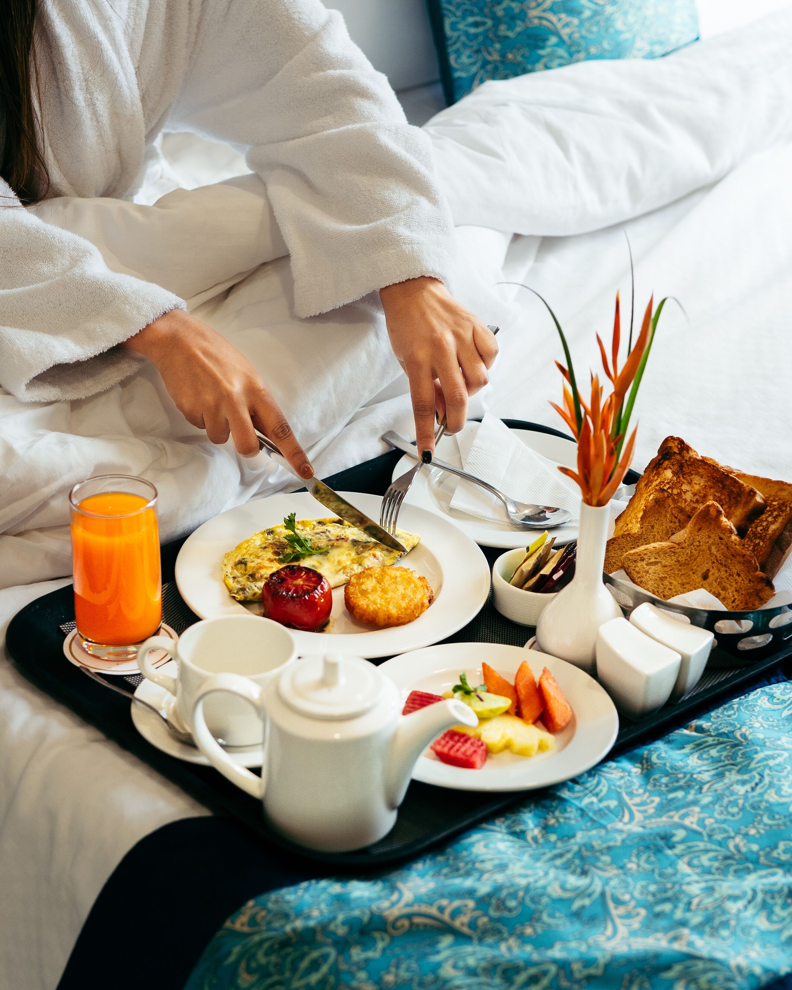 Bandeja de café da manhã com torradas, ovos, frutas e suco sobre uma cama com colcha azul estampada.