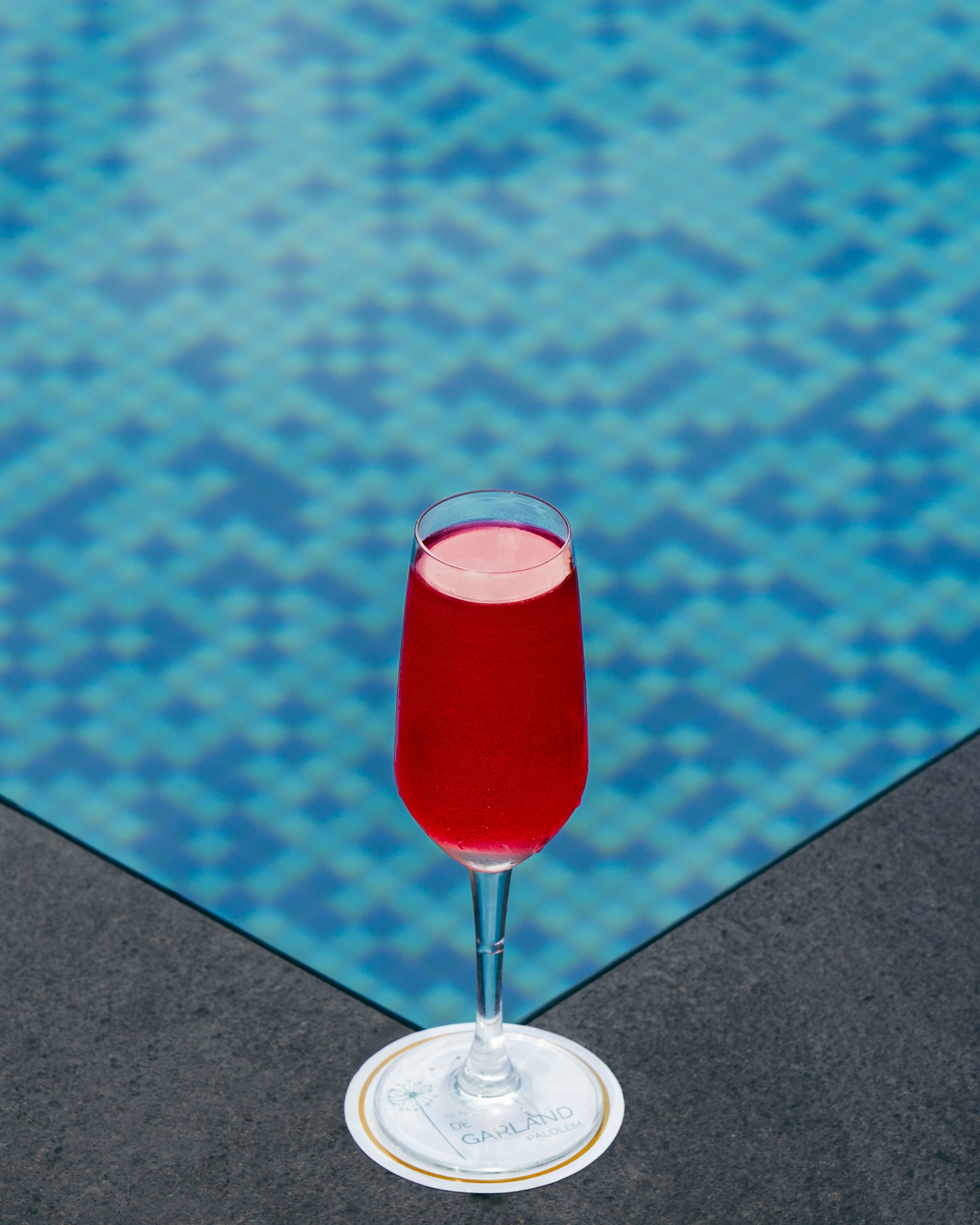 Um copo de bebida vermelha sobre uma mesa de vidro junto a uma piscina com piso de azulejos azuis estampados.