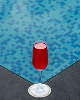 Um copo de bebida vermelha sobre uma mesa de vidro junto a uma piscina com piso de azulejos azuis estampados.