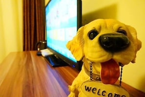 Uma figura de cão macia, com uma placa que diz 'Bem-vindo' na coleira, colocada sobre uma superfície de madeira com uma televisão ao fundo.