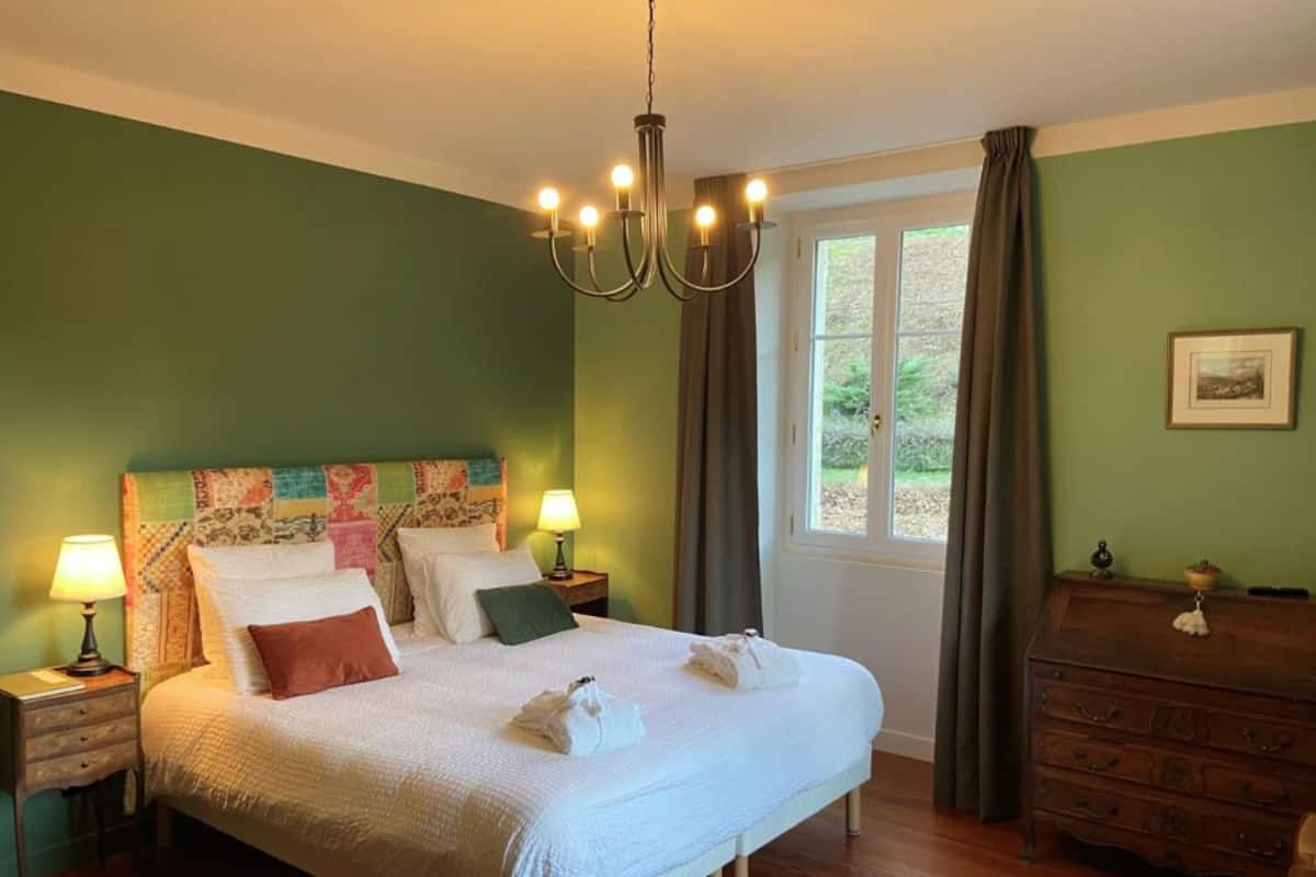 Quarto bem iluminado com uma cama grande, duas luminárias de cabeceira, cômoda de madeira e uma janela com cortinas.