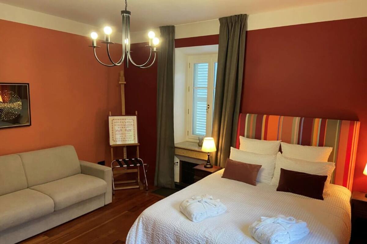 Quarto de hotel com cama, sofá, escrivaninha com luminária, espelho e lustre.