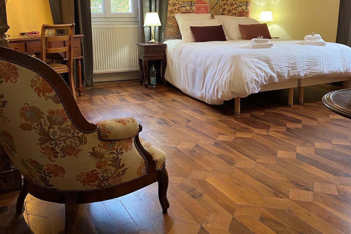 Quarto com cama, cadeira, mesa de cabeceira, abajur, lustre e janela com cortinas.
