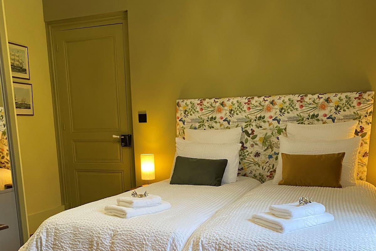 Uma cama de casal com lençóis brancos e duas almofadas, piso de madeira, porta verde e cabeceira estampada com flores.
