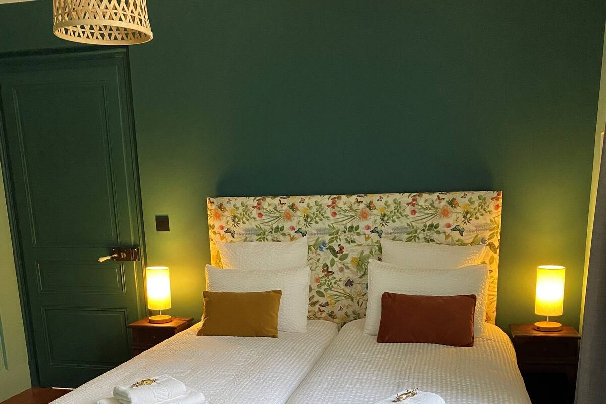 Uma cama de casal com roupa de cama branca, duas mesas de cabeceira com luminárias, um painel de cabeceira floral e uma porta verde.