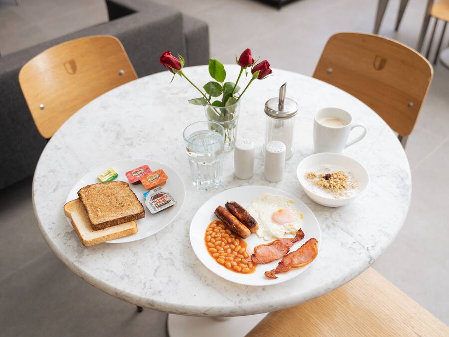 Mesa de café da manhã com torradas, feijão, bacon, ovos, linguiça e um vaso com rosas.