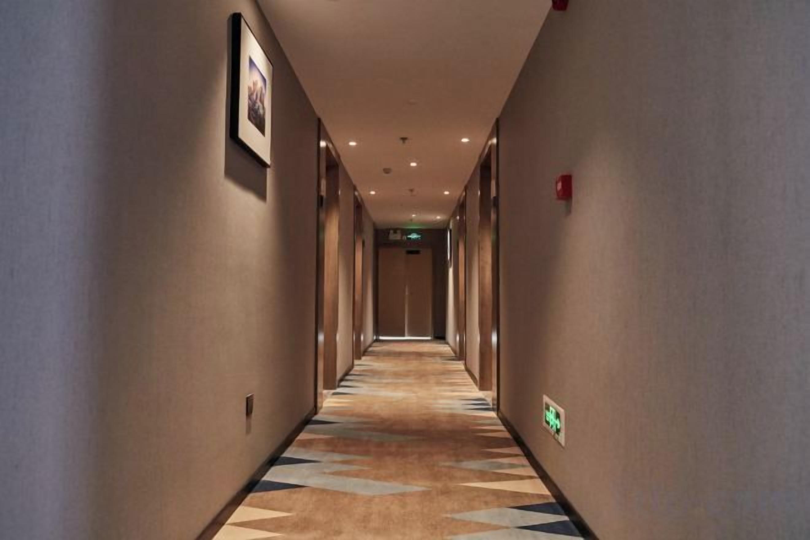 Corredor de hotel com piso carpetado, quadros nas paredes e um alarme de incêndio à direita.