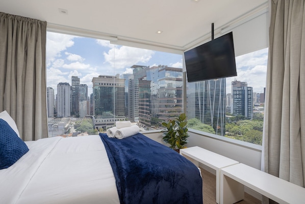 Quarto de hotel com uma cama grande, televisão, um banco e vista para a paisagem urbana.