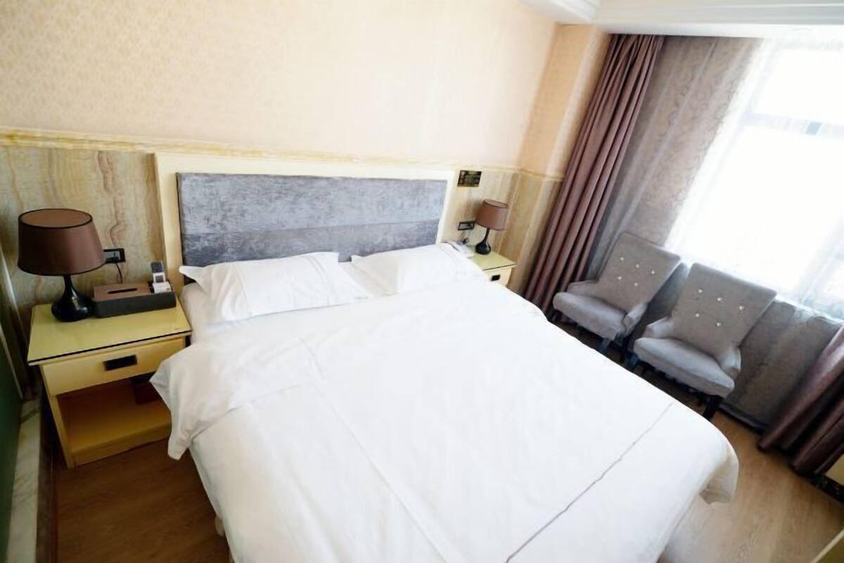 Quarto de hotel com uma cama grande, duas mesas de cabeceira com luminárias, uma cadeira e uma janela com cortinas.