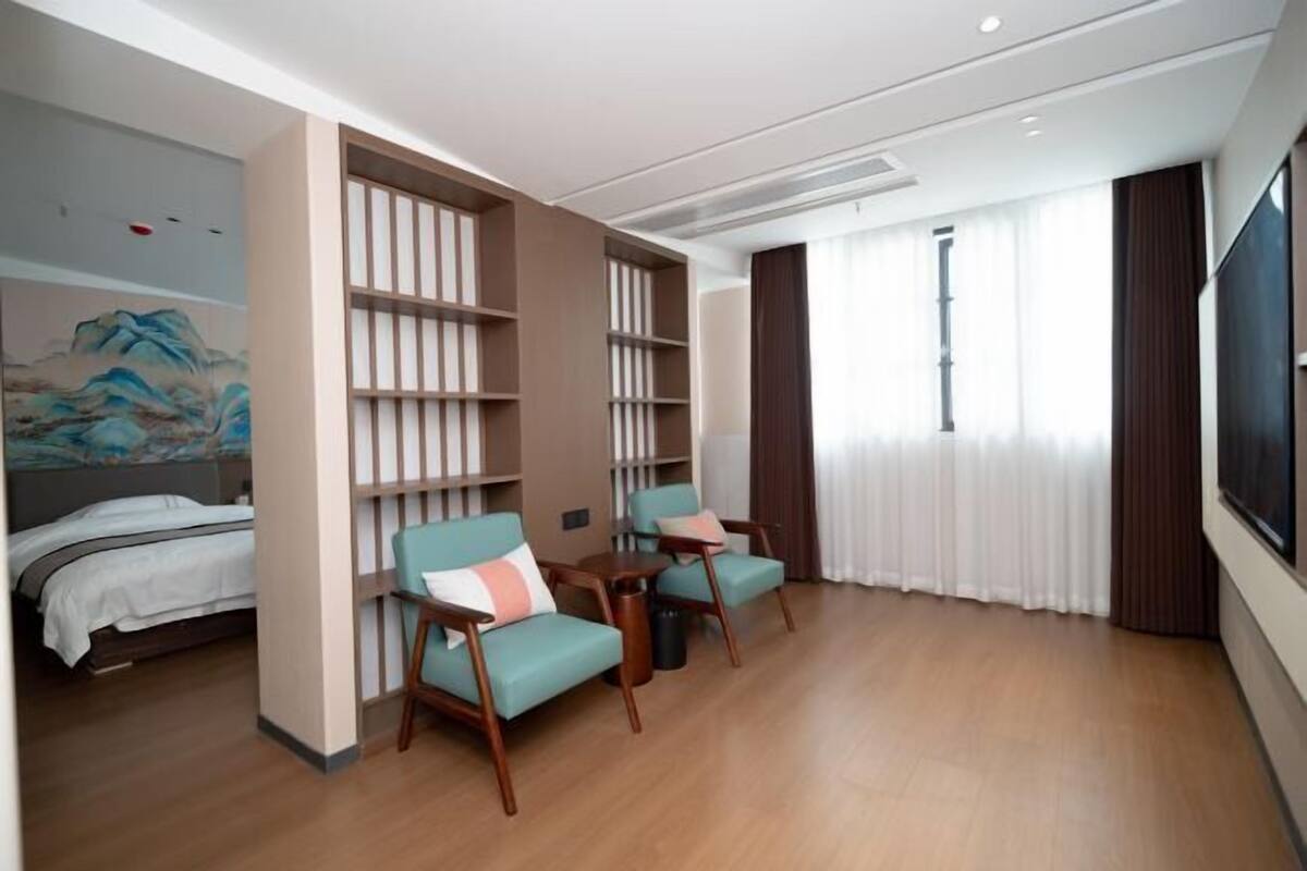 Quarto de hotel com cama, duas poltronas, uma mesa pequena, estante de livros e uma janela grande com cortinas.