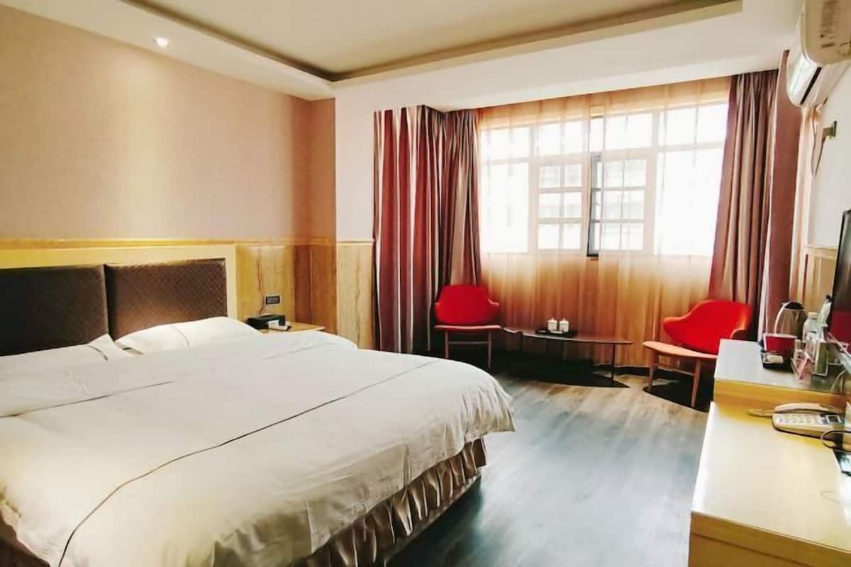 Quarto de hotel com uma cama grande, uma escrivaninha com telefone, duas cadeiras vermelhas e uma janela com cortinas.