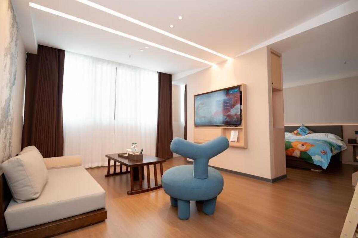 Quarto de hotel moderno com sofá, uma cadeira azul exclusiva, mesa de jantar e cama com colcha colorida.