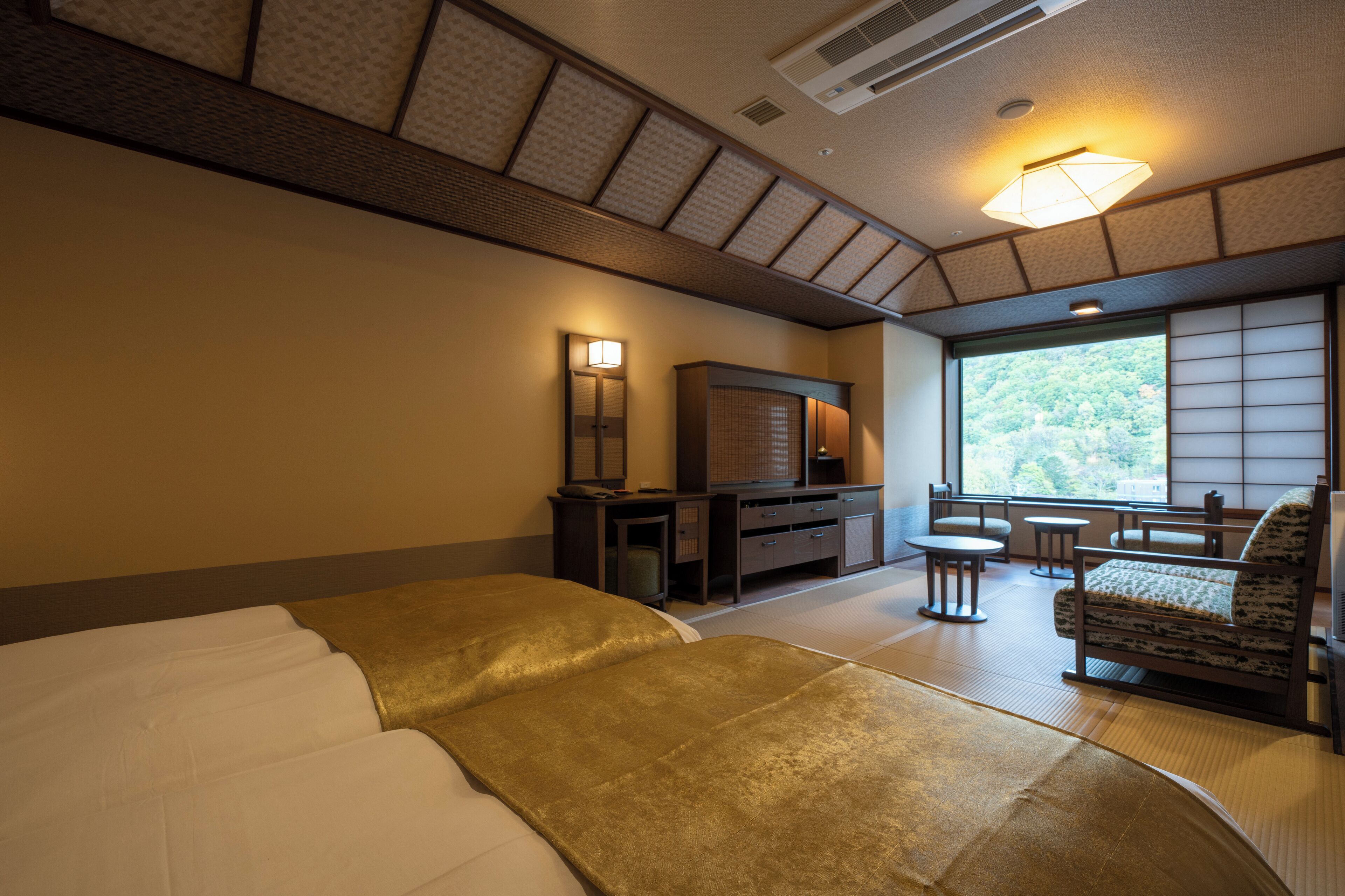 Quarto de hotel em estilo japonês tradicional, com piso de tatame, cama com detalhes dourados, escrivaninha com luminária e vista para o verde através de grandes janelas.