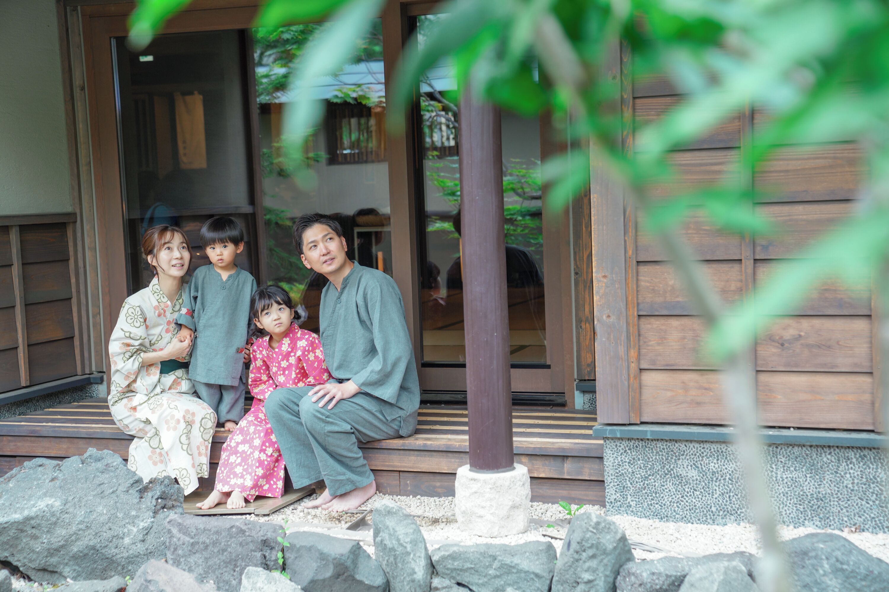 Uma família sentada em um alpendre de madeira com arquitetura japonesa tradicional, incluindo portas corrediças e um caminho de pedra.