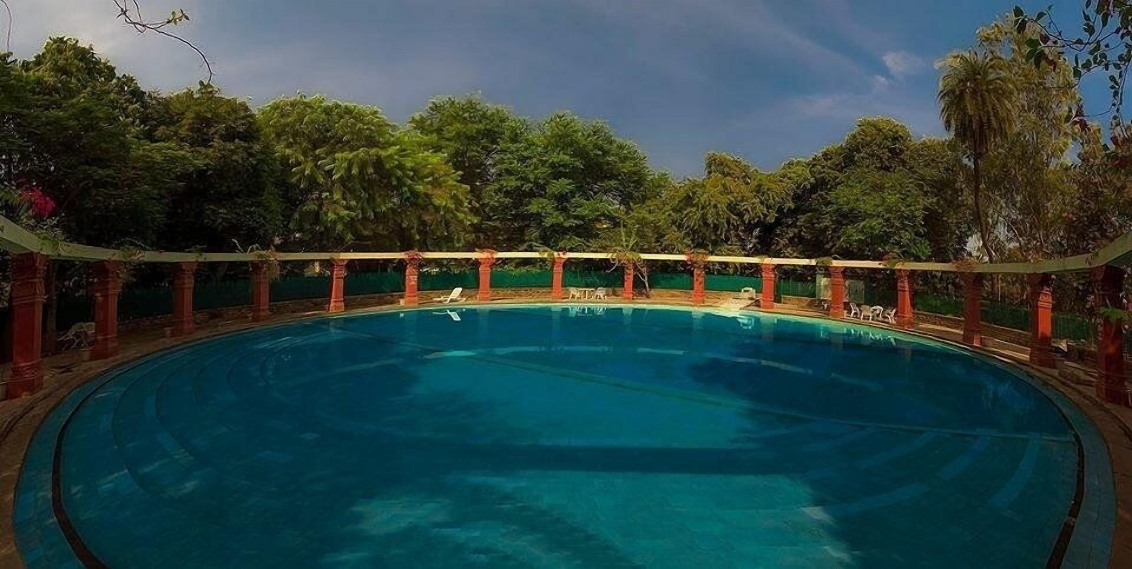 Uma grande piscina em formato oval, cercada por uma vedação vermelha e branca, com árvores e vegetação ao fundo.