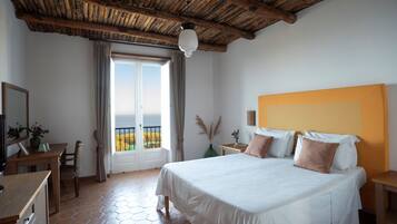Quarto com uma cama grande, teto de madeira, uma escrivaninha com cadeira, janela com cortinas e vista para o exterior.