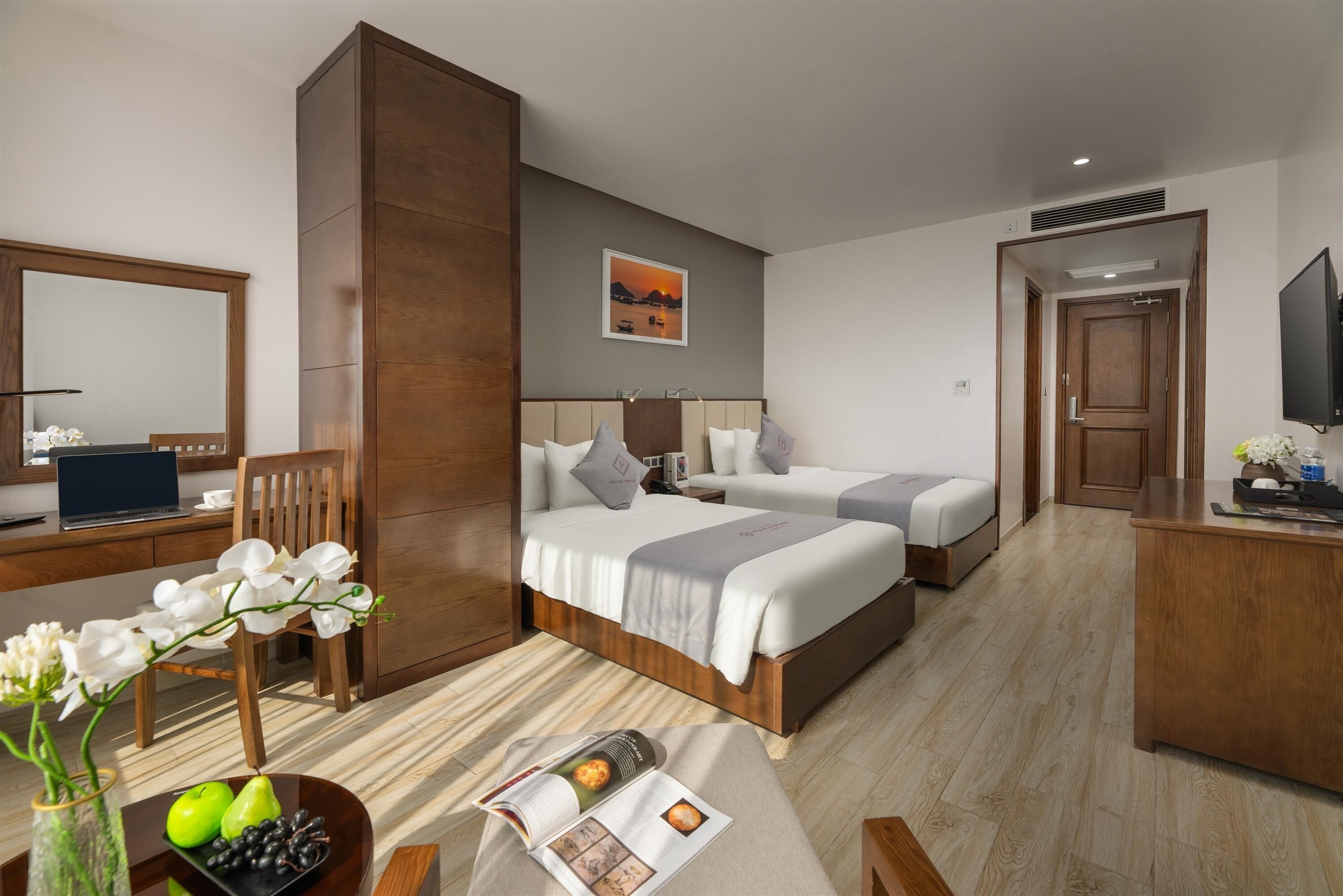 Quarto de hotel com duas camas, uma escrivaninha com um laptop, um espelho e um vaso com flores.