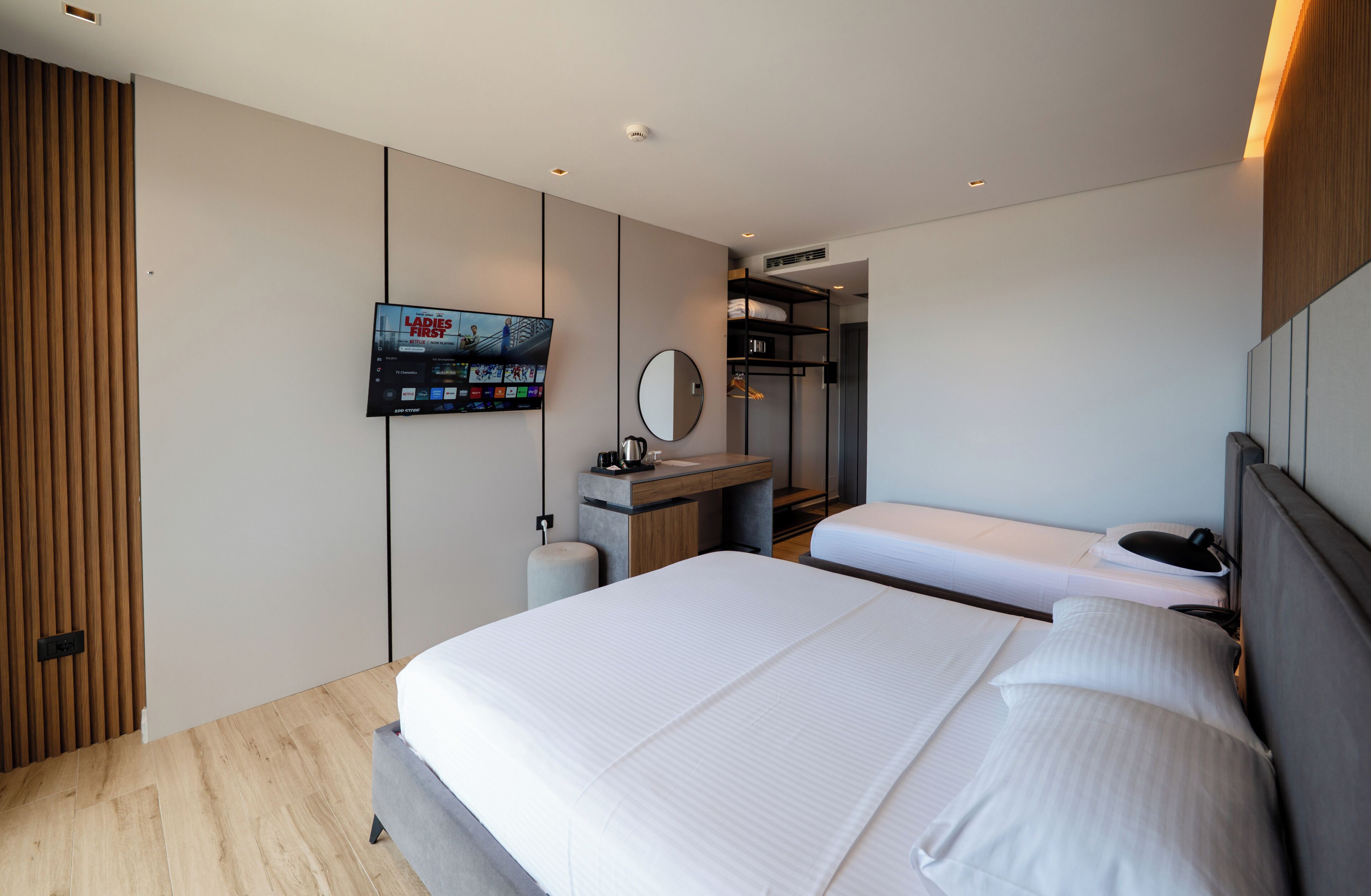 Quarto de hotel moderno com uma cama grande, uma cama menor, TV na parede, escrivaninha com cadeira e guarda-roupa.