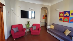 Sala de estar com televisão de tela plana, duas poltronas vermelhas, um sofá cinza e obras de arte coloridas nas paredes.