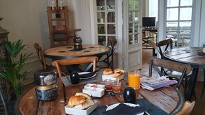 Uma área de refeições com mesas e cadeiras de madeira, um vaso decorativo e uma mesa de café da manhã com croissants e suco de laranja.