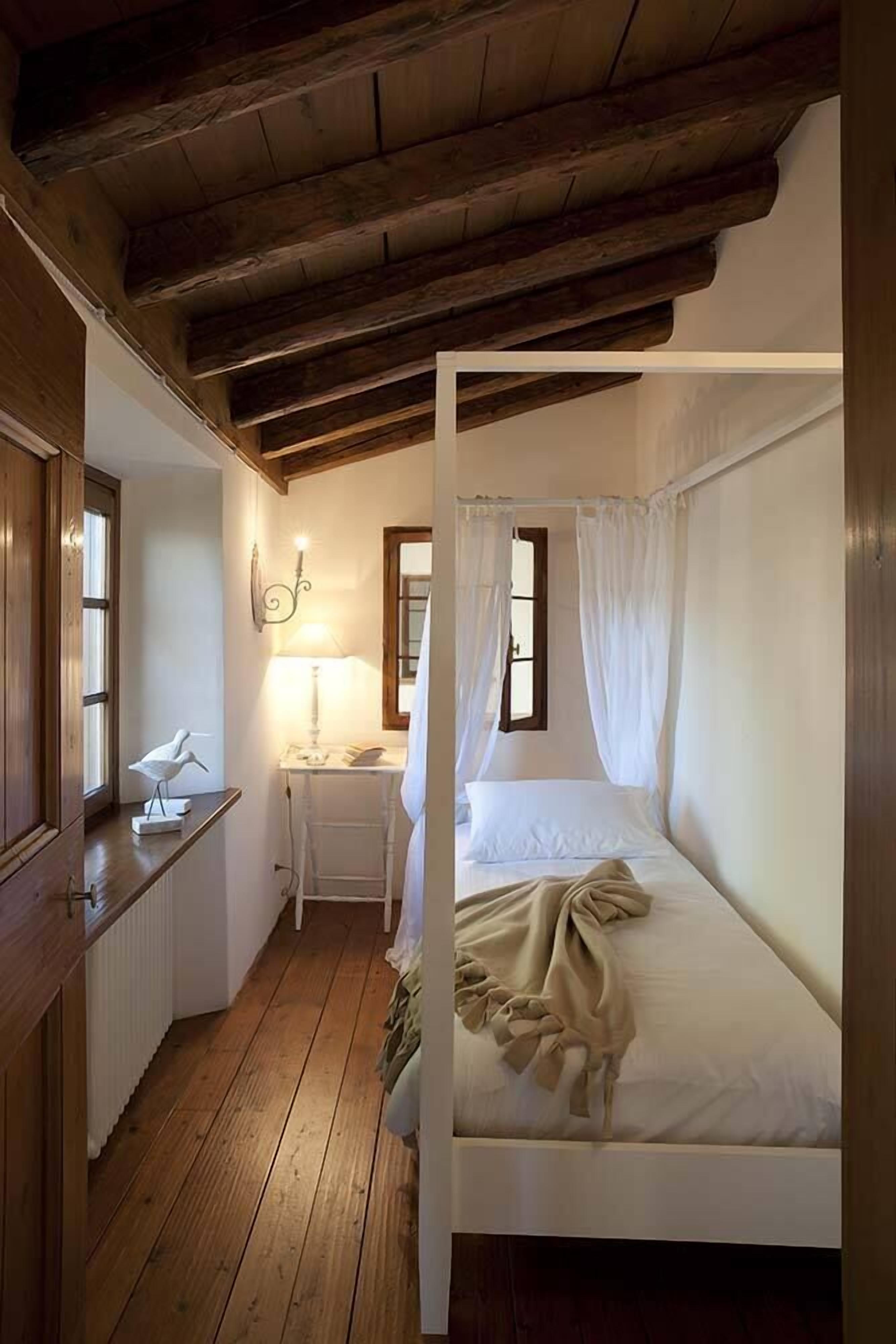 Um quarto com teto de madeira, uma cama com dossel, uma mesa de cabeceira com abajur e uma janela com cortinas.