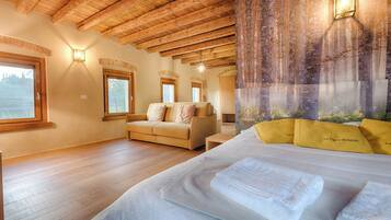 Quarto com teto de madeira, cama com lençóis brancos, sofá com almofadas amarelas e vista para árvores pela janela.