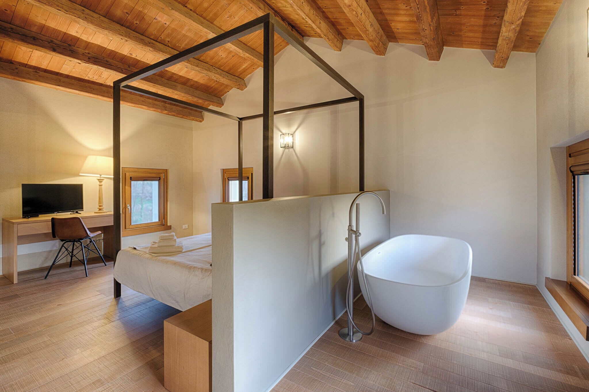 Quarto moderno com teto de madeira, uma cama grande, uma escrivaninha com cadeira, televisão e uma banheira ampla.