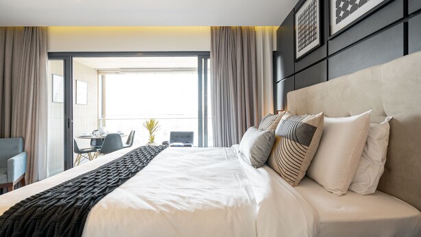Quarto de hotel moderno com uma cama grande, um sofá e uma área de refeições visível através de uma porta aberta.