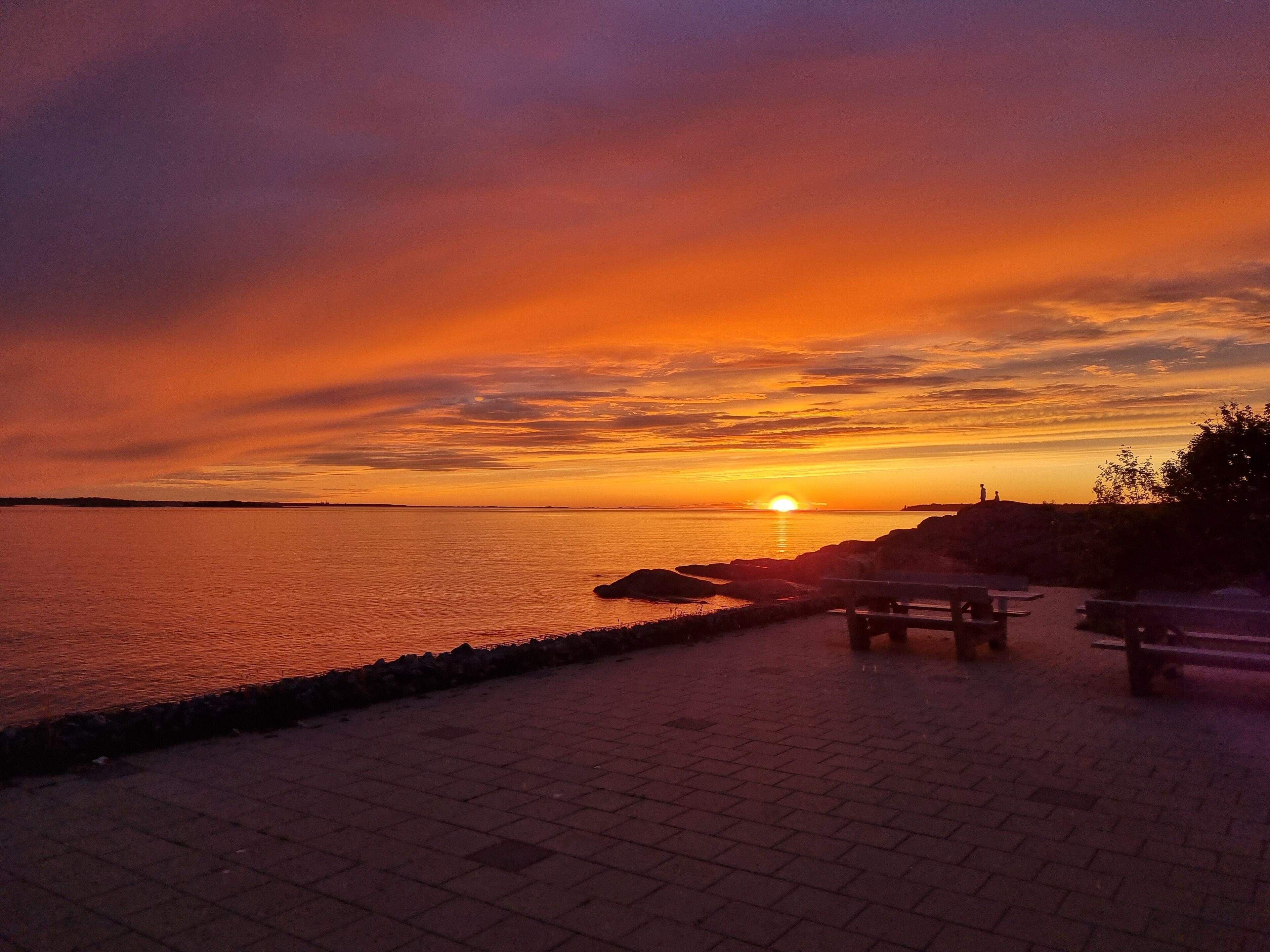 Um pôr do sol sobre águas calmas, com um calçadão pavimentado e bancos.