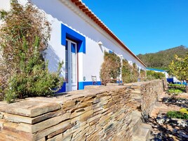 Um edifício tradicional com parede de pedra e porta azul, cercado por vegetação e céu claro.