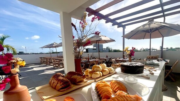 Buffet ao ar livre com diversos alimentos, incluindo pão, doces e frutas fatiadas, sob área coberta com guarda-sóis.