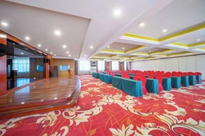 Uma sala de conferências com palco, fileiras de mesas e cadeiras vermelhas, e um tapete estampado.