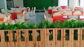 Uma sala de conferências com fileiras de cadeiras brancas com fitas laranja, uma mesa vermelha e uma parede verde com plantas.