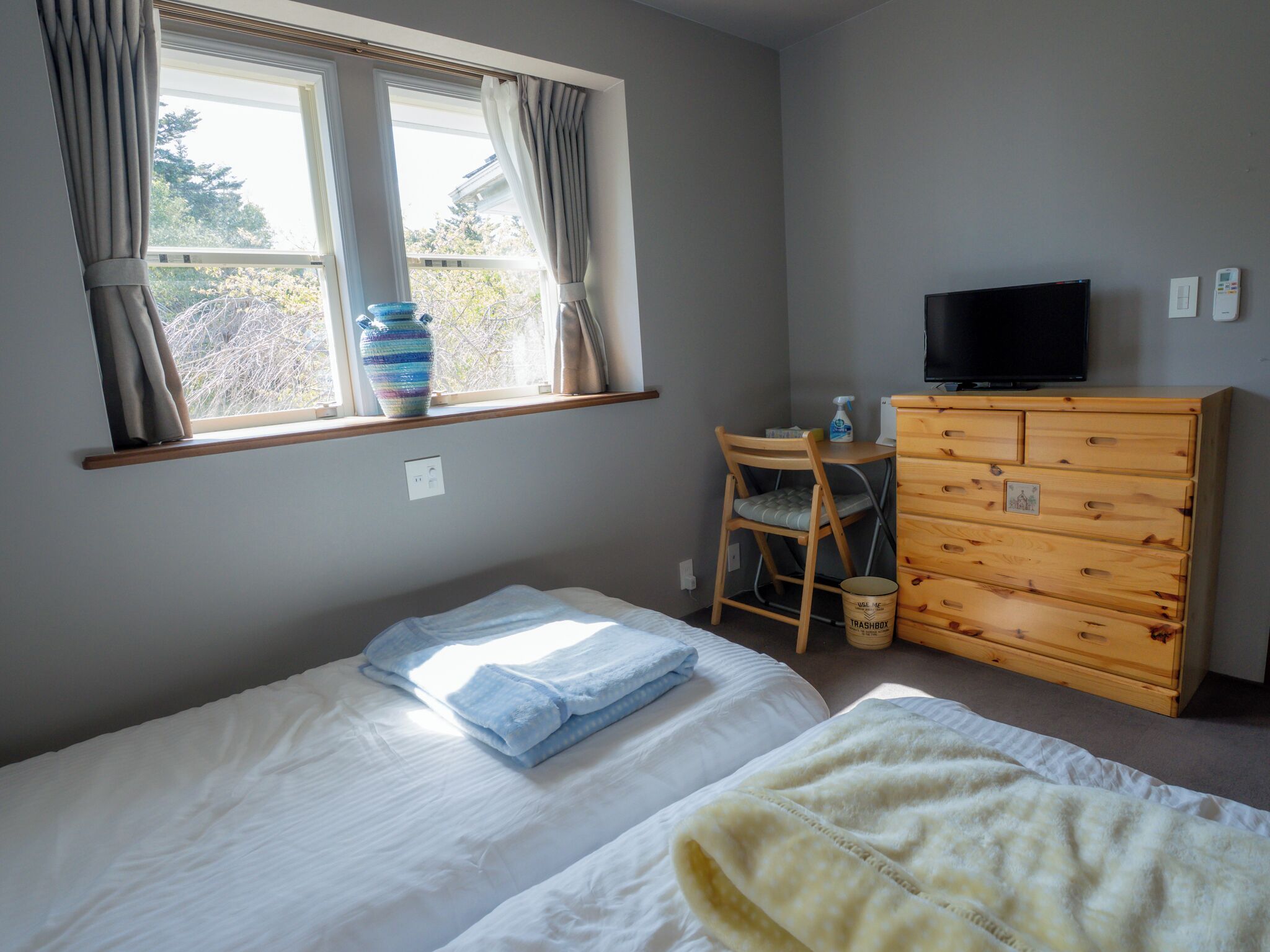 Quarto com duas camas, televisão, penteadeira de madeira, uma cadeira e janela com cortinas.