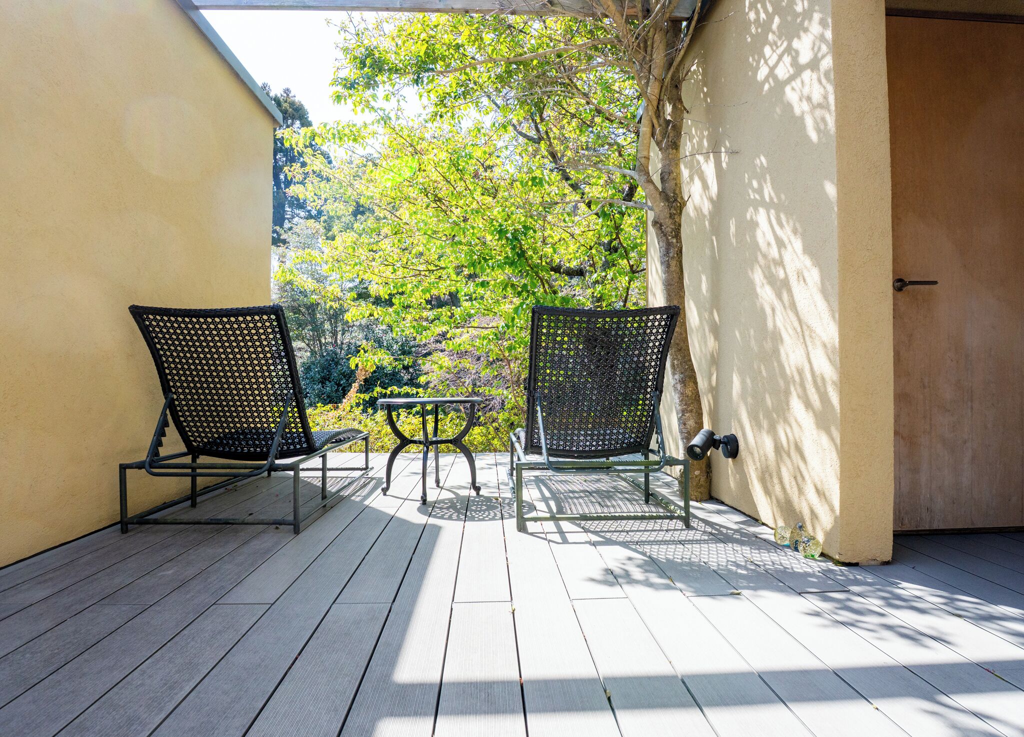 Um pátio externo com duas cadeiras, uma mesa, um deck de madeira e vista para árvores e um edifício.
