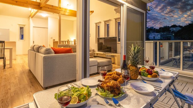 Uma mesa de jantar posta com comida e bebidas, um sofá e uma TV numa sala de estar moderna com vigas de madeira e varanda com vista para a cidade.