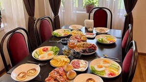 Mesa de jantar posta para seis pessoas com diversos itens de café da manhã, cercada por cadeiras estofadas vermelhas, e vista para o verde através de grandes janelas.
