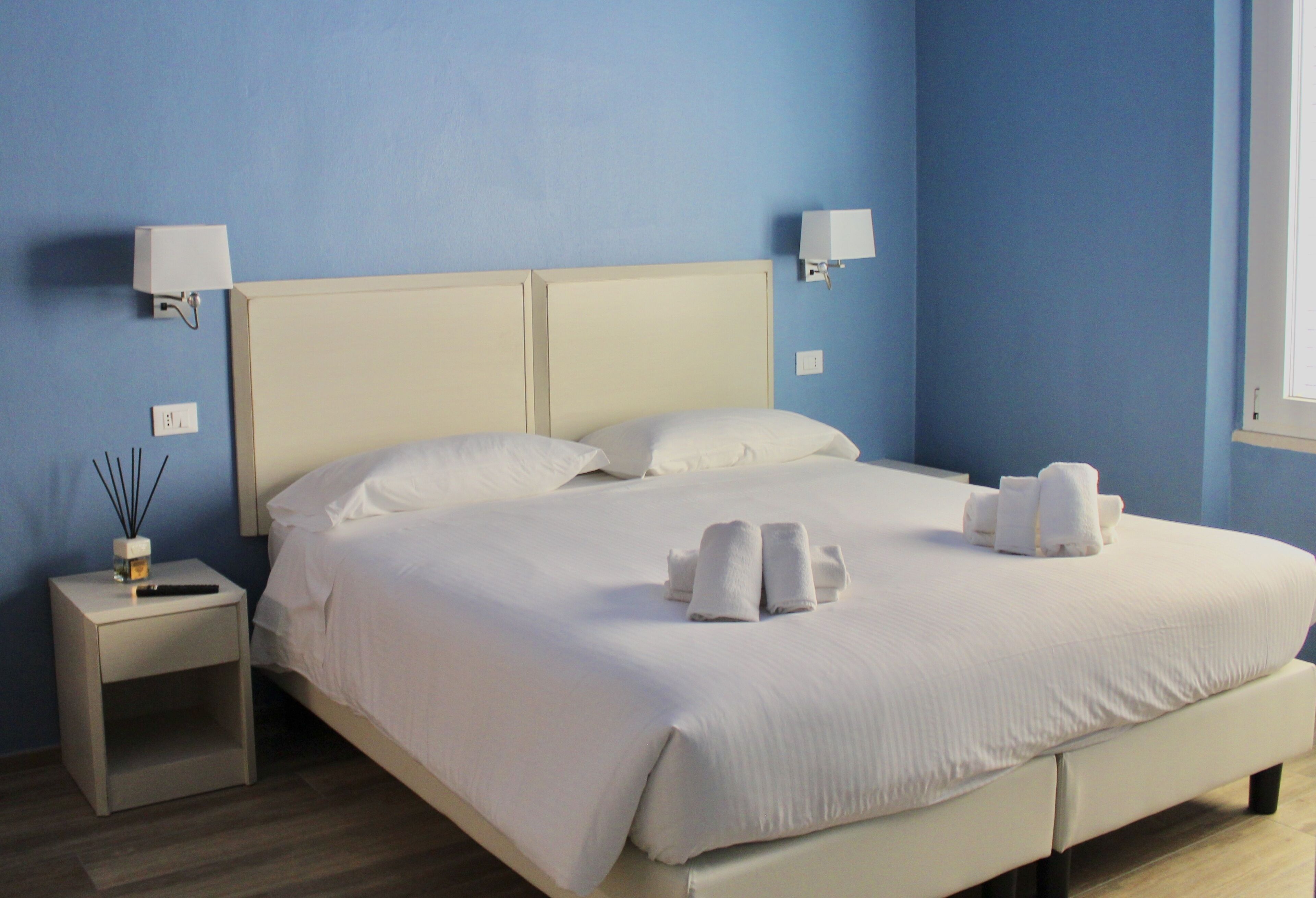 Cama bem arrumada com roupa de cama e toalhas brancas, mesa de cabeceira com abajur e luminária na parede.