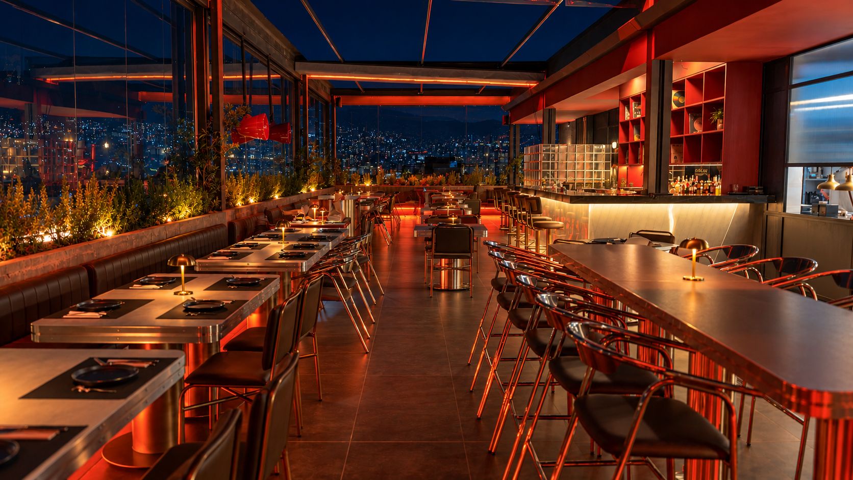 Um bar no terraço com mesas, cadeiras e balcão, com vista para a cidade à noite.