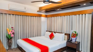 Quarto de hotel com uma cama grande, cabeceira de madeira, ventilador de teto, aparelho de ar condicionado, mesa de cabeceira com telefone e um vaso de flores, além de uma planta decorativa.