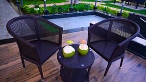 Uma área de pátio com cadeiras de vime e uma pequena mesa, dois cocos sobre a mesa, e vista para o jardim.