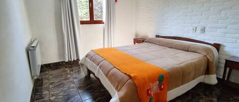 Um quarto com cama, janela com cortinas, radiador e parede de tijolos brancos.