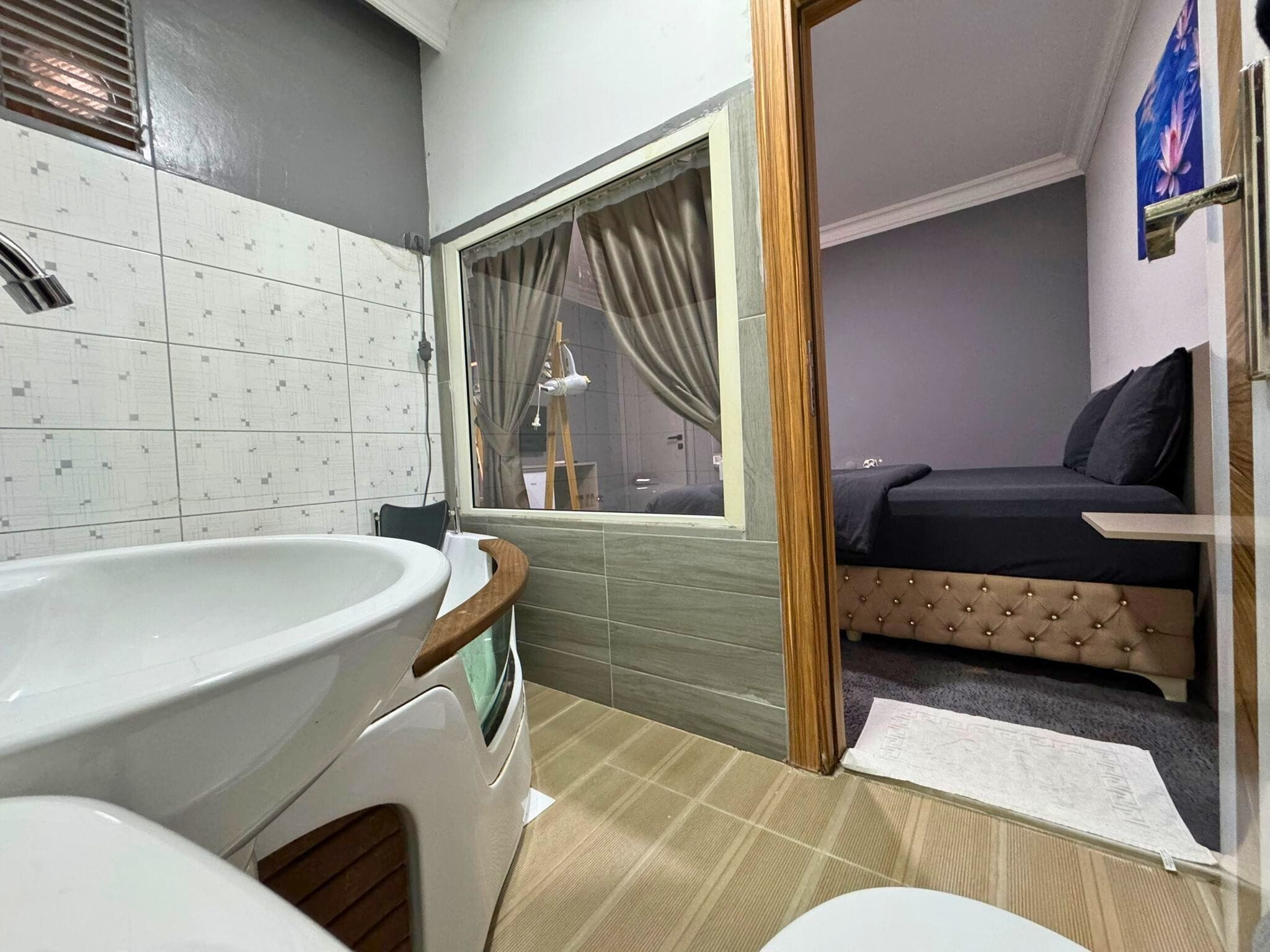 Quarto de hotel compacto com uma pia branca grande, uma cama pequena com cabeceira estofada, uma janela com cortinas e uma parede revestida de azulejos.