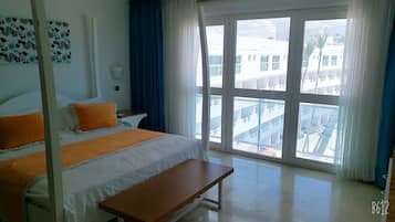 Quarto de hotel com cama, um banquinho, mesa de cabeceira e janela com vista para prédios.