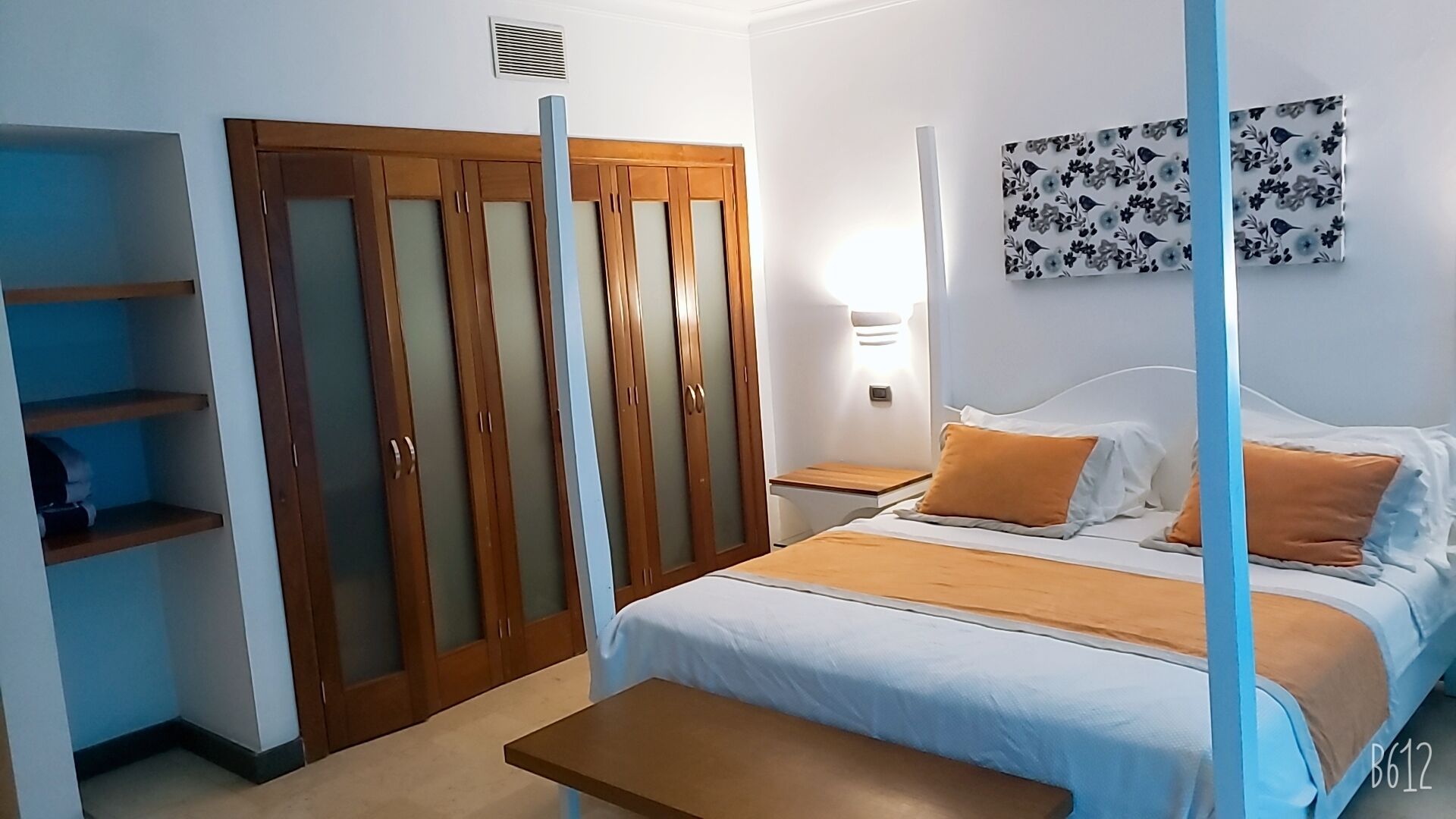 Quarto de hotel com cama, mesa de cabeceira, guarda-roupa e estante.