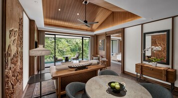 Quarto de hotel moderno com uma janela grande que oferece vista para vegetação exuberante, um ventilador de teto e uma área de jantar com mesa e cadeiras redondas.