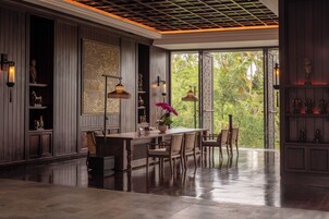 Um quarto espaçoso com piso de madeira, uma mesa de jantar central com cadeiras, uma janela grande com vista para o verde e arandelas decorativas na parede.