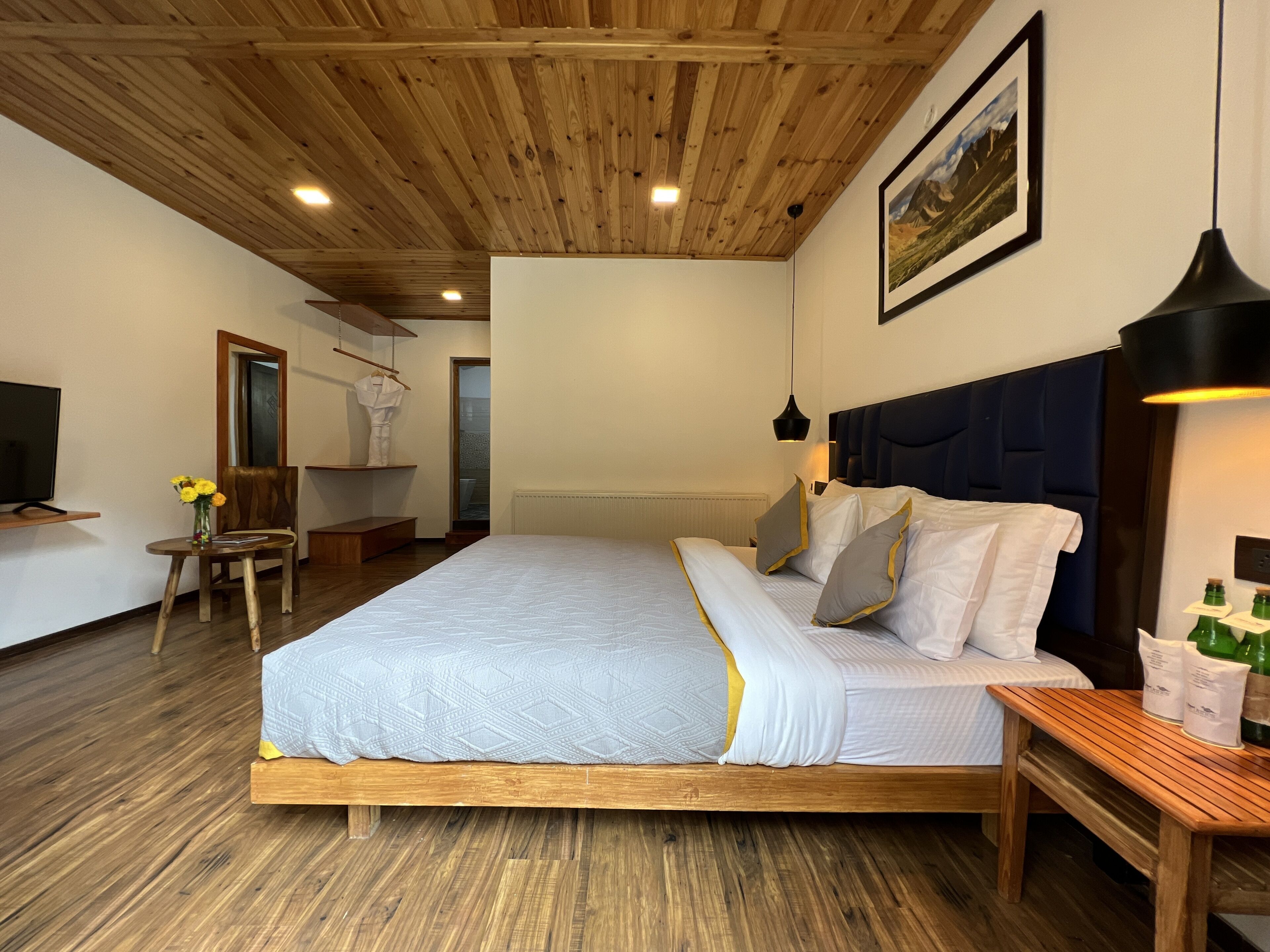 Quarto moderno com teto de madeira, cama grande com roupa de cama branca, mesa de cabeceira com abajur e pequena mesa redonda com um vaso de flores.